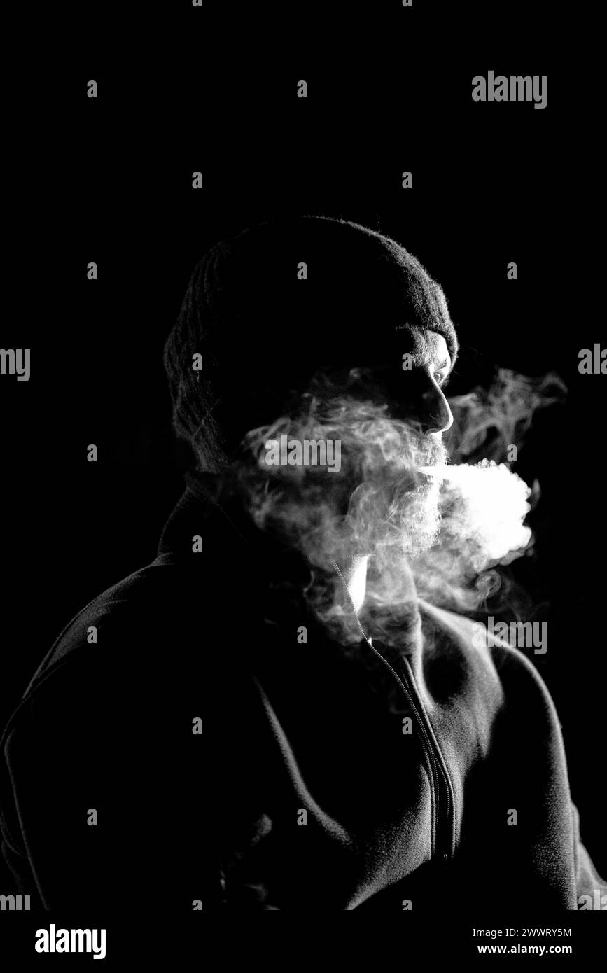 A dark grayscale shot of a man in a hat and jacket smoking a cigarette ...