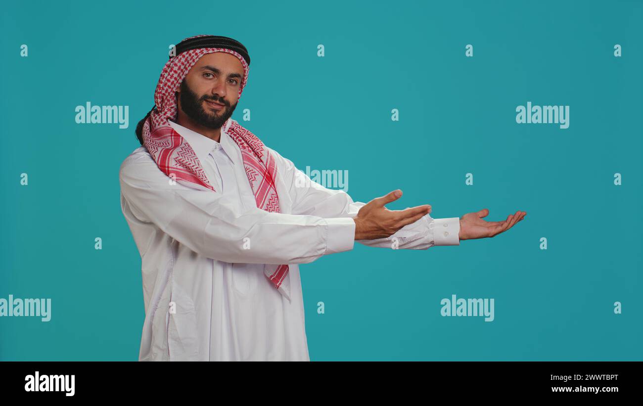 Muslim adult in traditional gown and ghutra presenting sides with hands ...