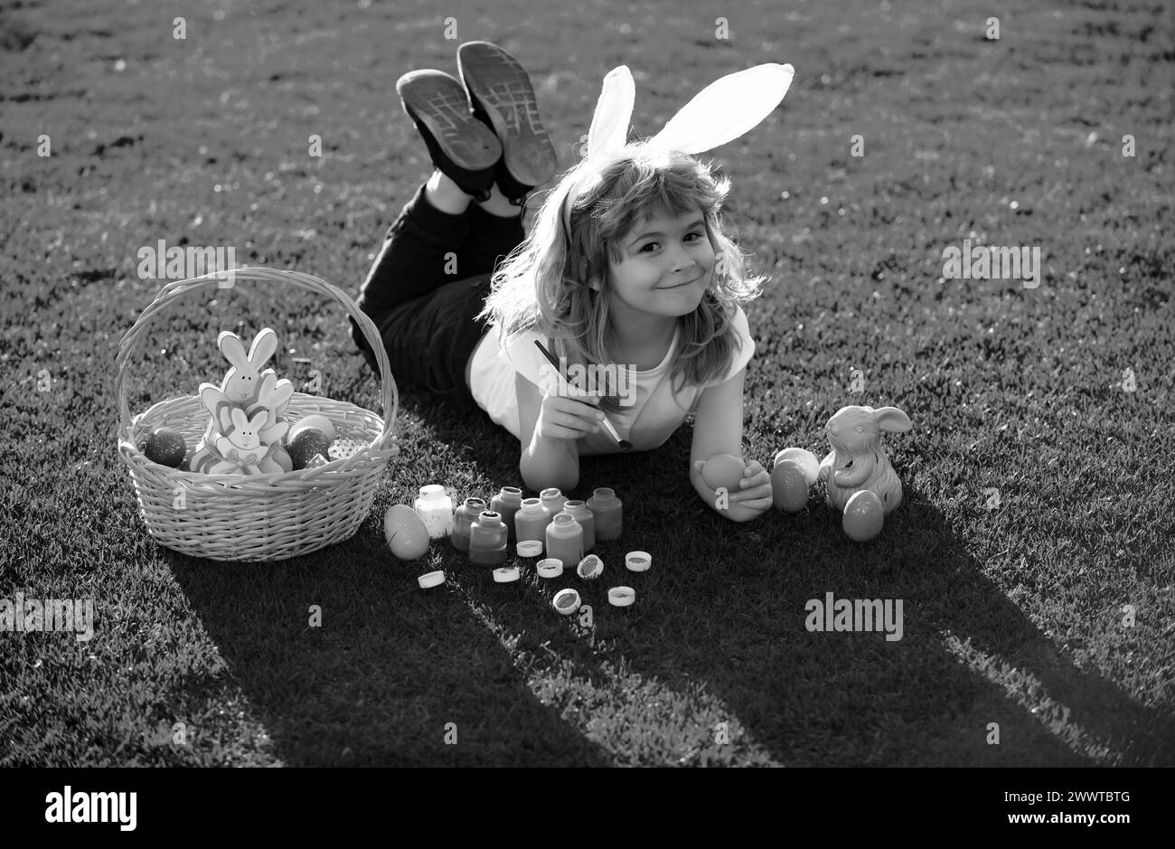 Child with easter eggs and bunny ears laying on grass painting eggs ...