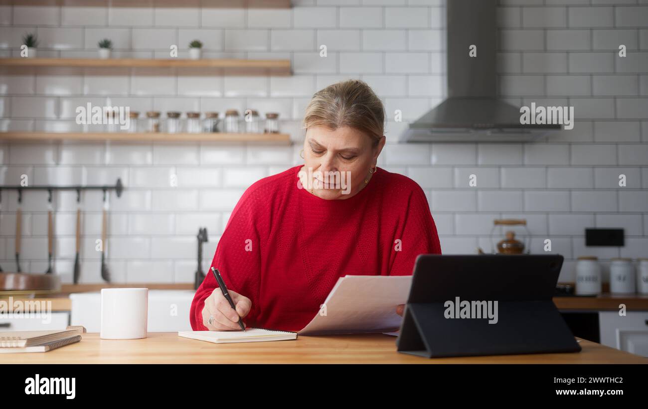 Elderly business woman sit in kitchen using tablet device, check ...