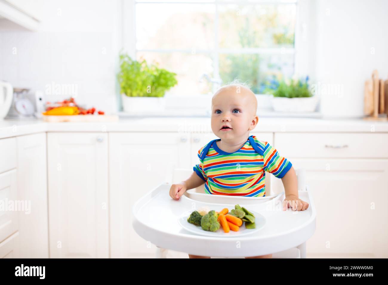 Cute baby eating vegetables in white kitchen. Infant weaning. Little ...