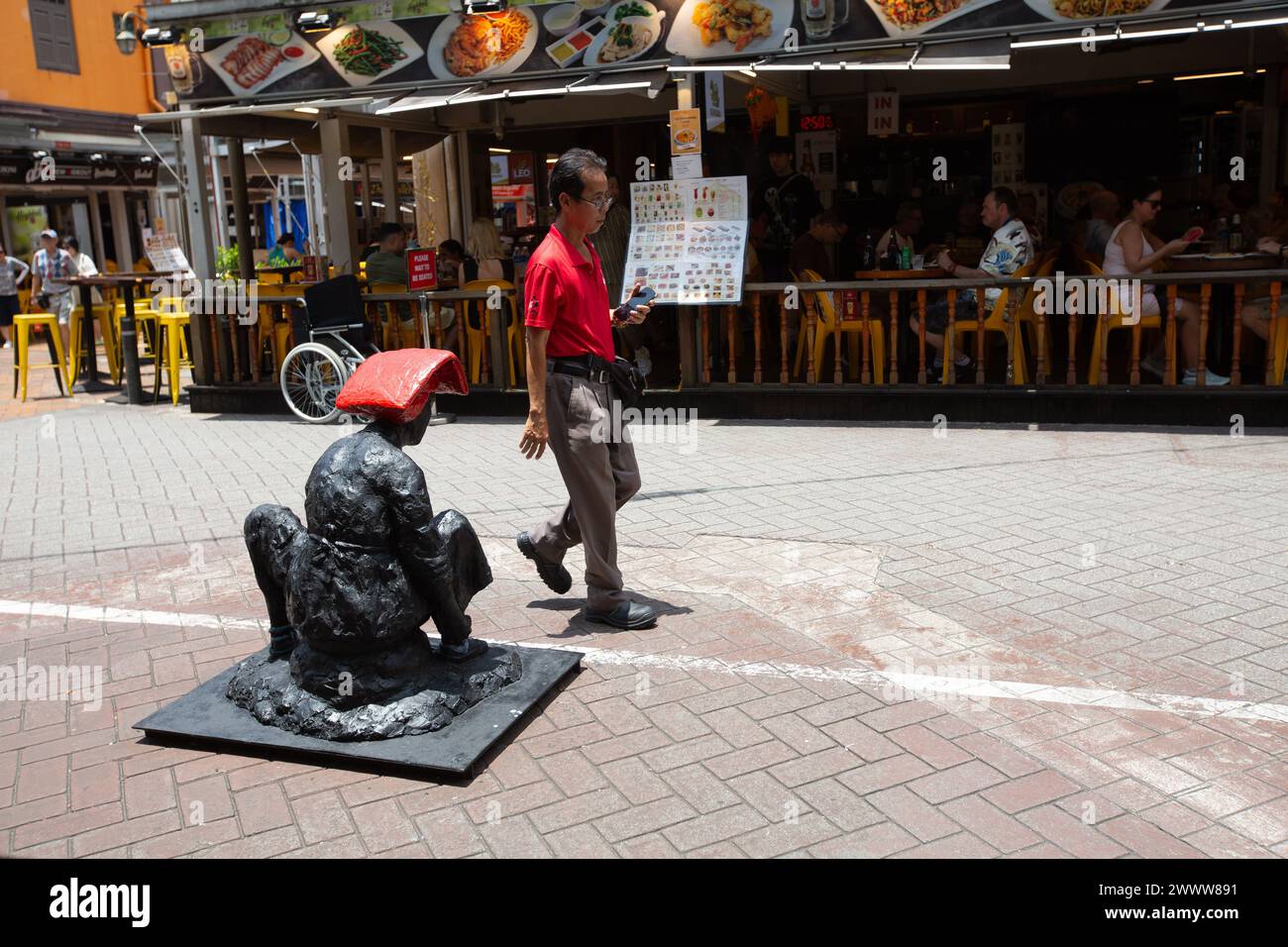 Chinese man in red shirt under the hot sun walks past a Samsui woman ...