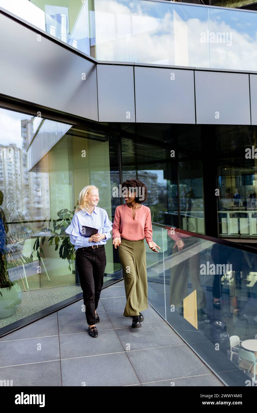 Two colleagues laugh together, one holding a tablet in a sunny, glass ...