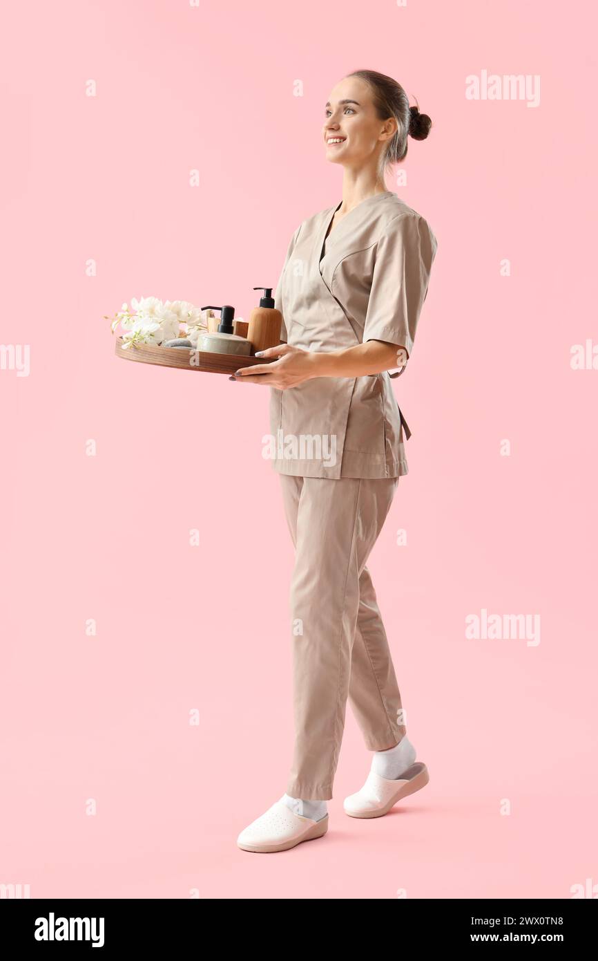 Portrait of female massage therapist holding tray with spa supplies and flowers on pink ...