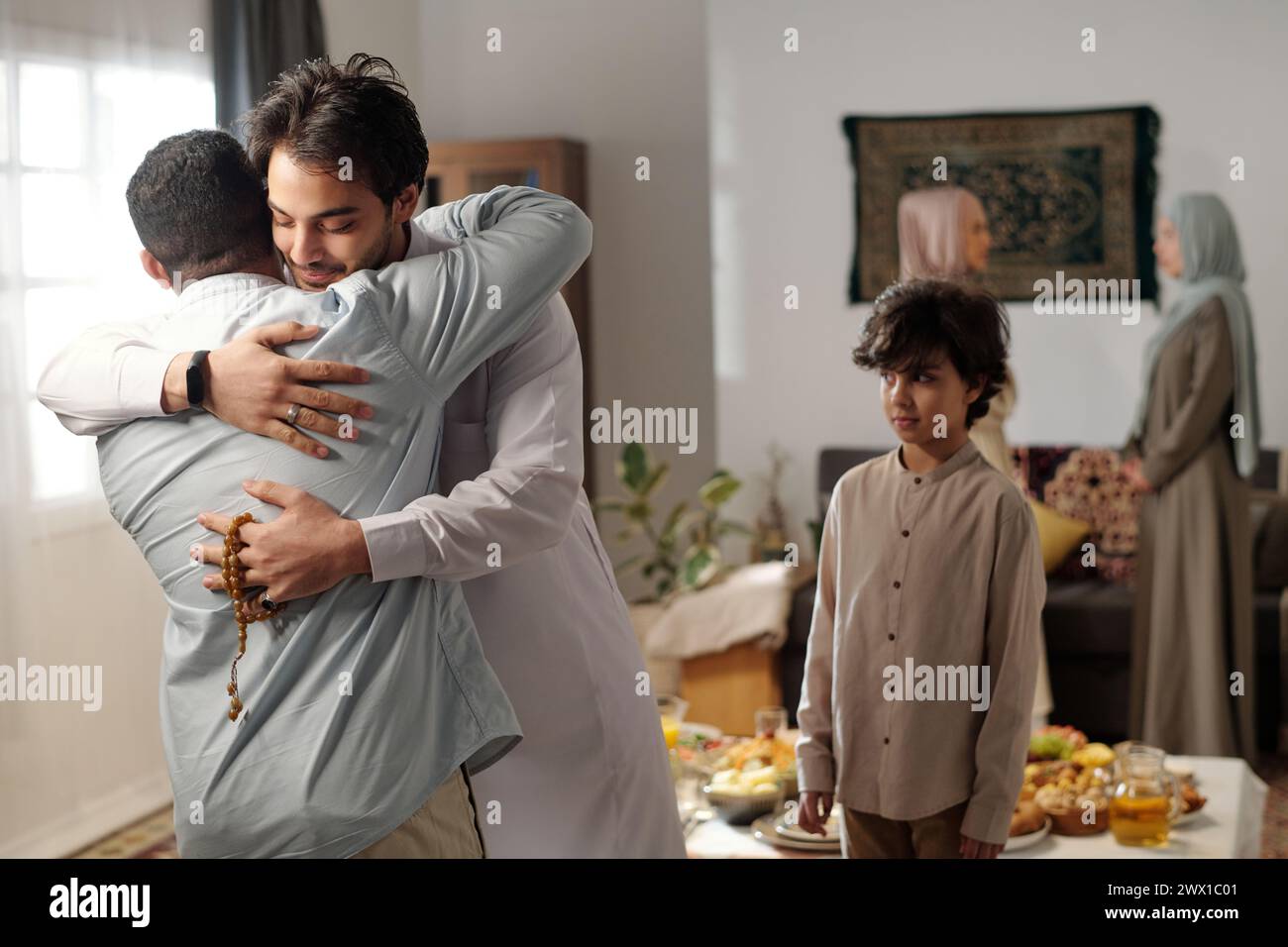 Medium shot of young Muslim man wearing thobe greeting male relative with hug on Eid al-Fitr ...