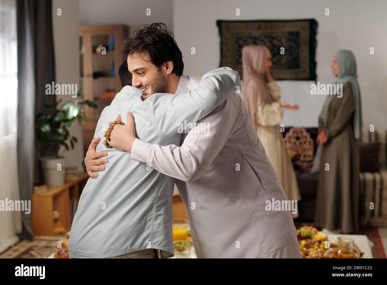 Medium side view of two Muslim men greeting each other with hug on Eid Al-Fitr, two women ...