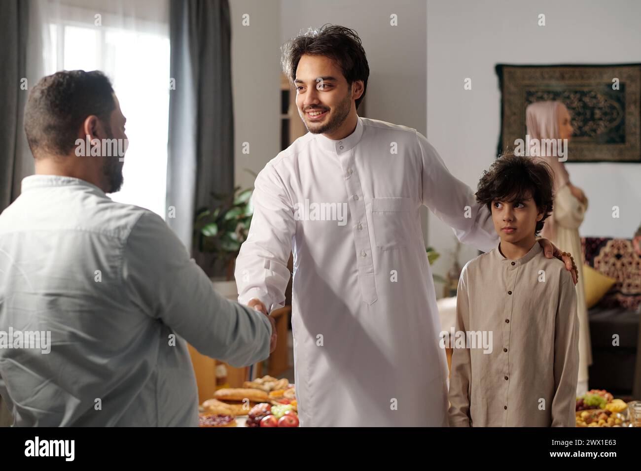 Medium shot of young Muslim man and his son wearing thobes greeting ...