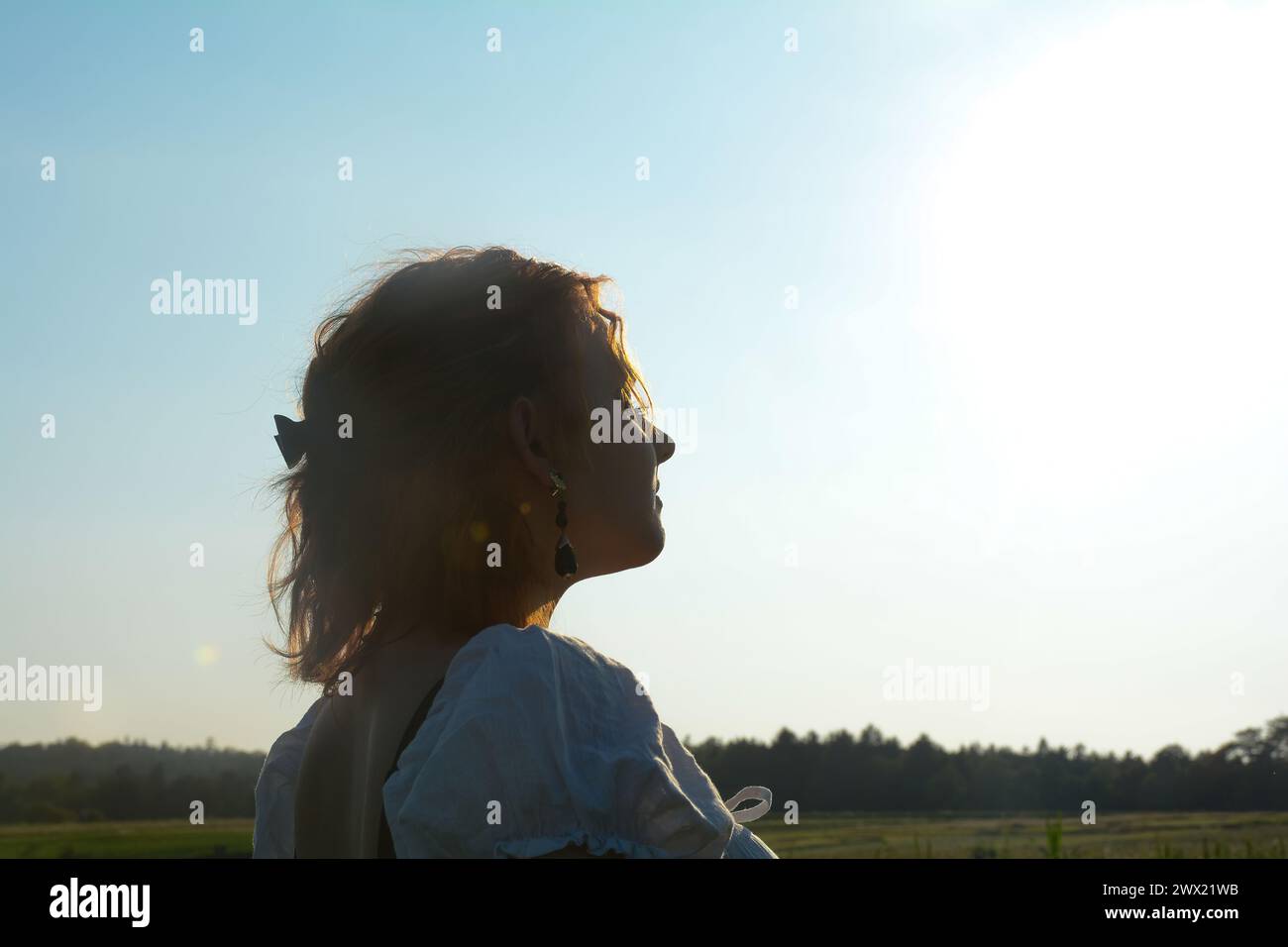 Profile of a pretty young woman, facing the sun and standing in green ...