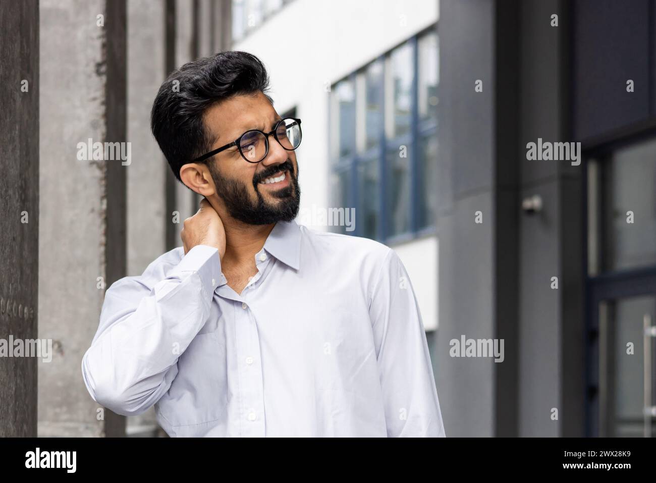 Tired young Indian male office worker standing outside near building and holding neck, giving ...