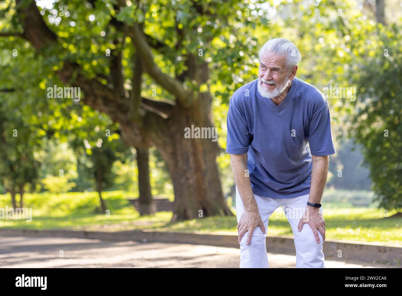 Tired senior gray-haired man is standing bent over in the park and ...