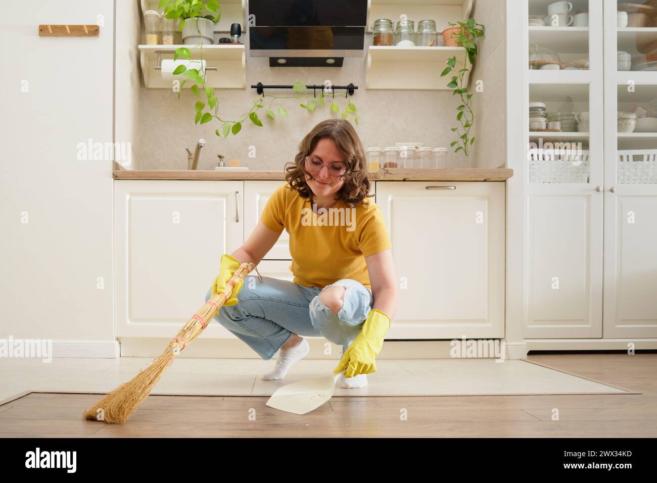 A woman sweeps the floor with a broom and dustpan in her home kitchen ...