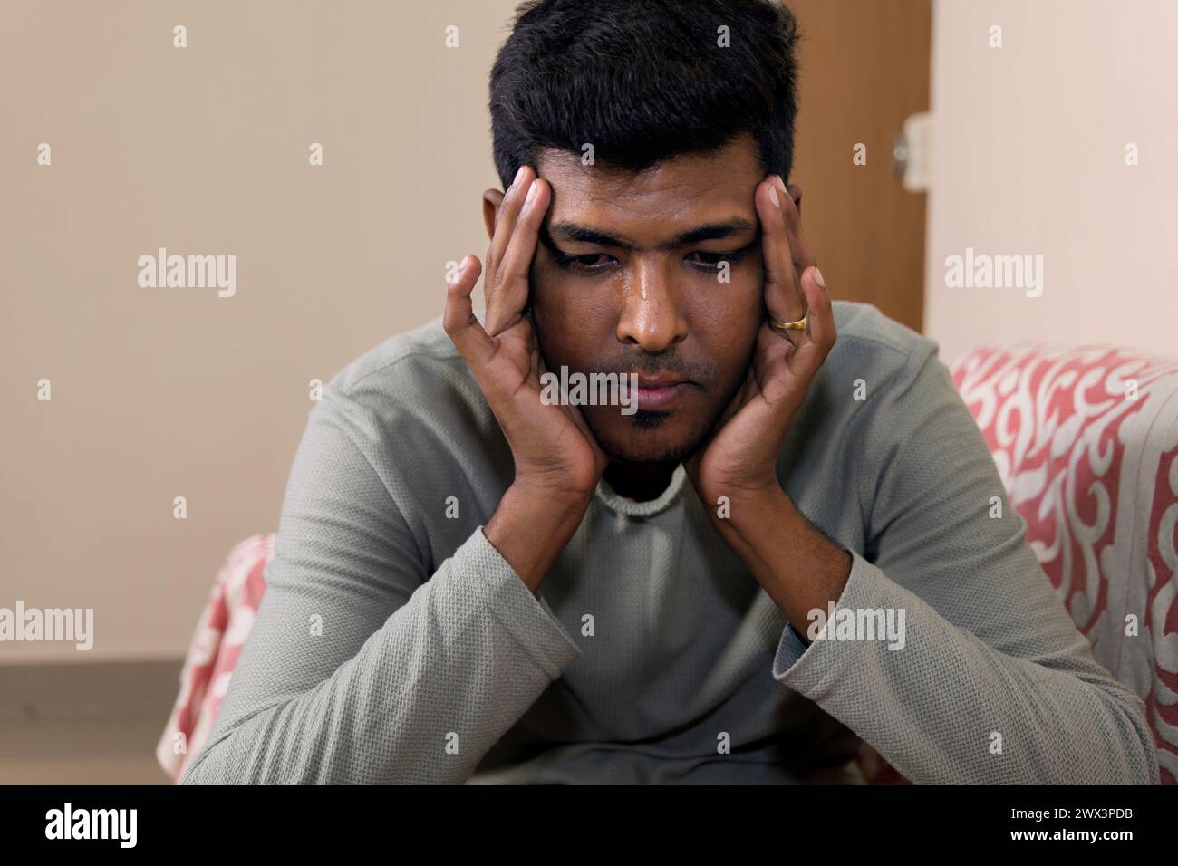 A young Indian man sits alone on a couch in his living room, lost in ...