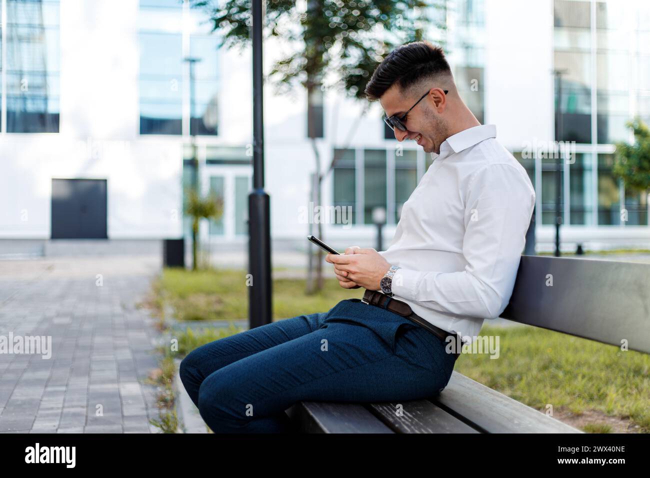 A man is sitting on a bench outdoors, engrossed in his cell phone. He ...
