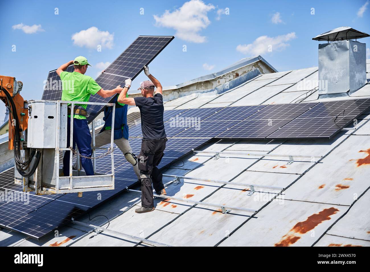 Workers lifting up photovoltaic solar panel on metal rooftop of house with assistance of crane ...