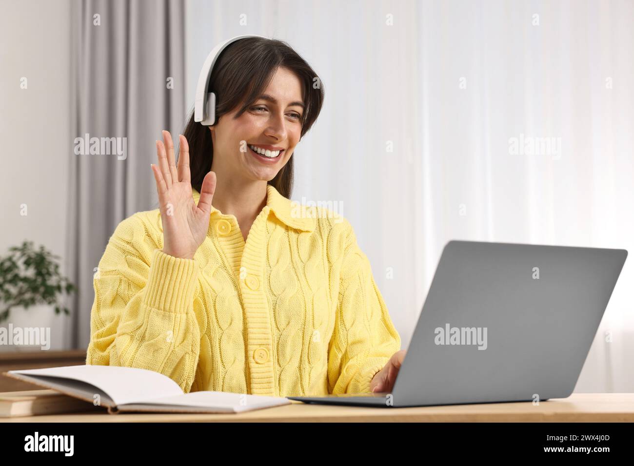 Happy woman waving hello during video chat via laptop at table indoors ...