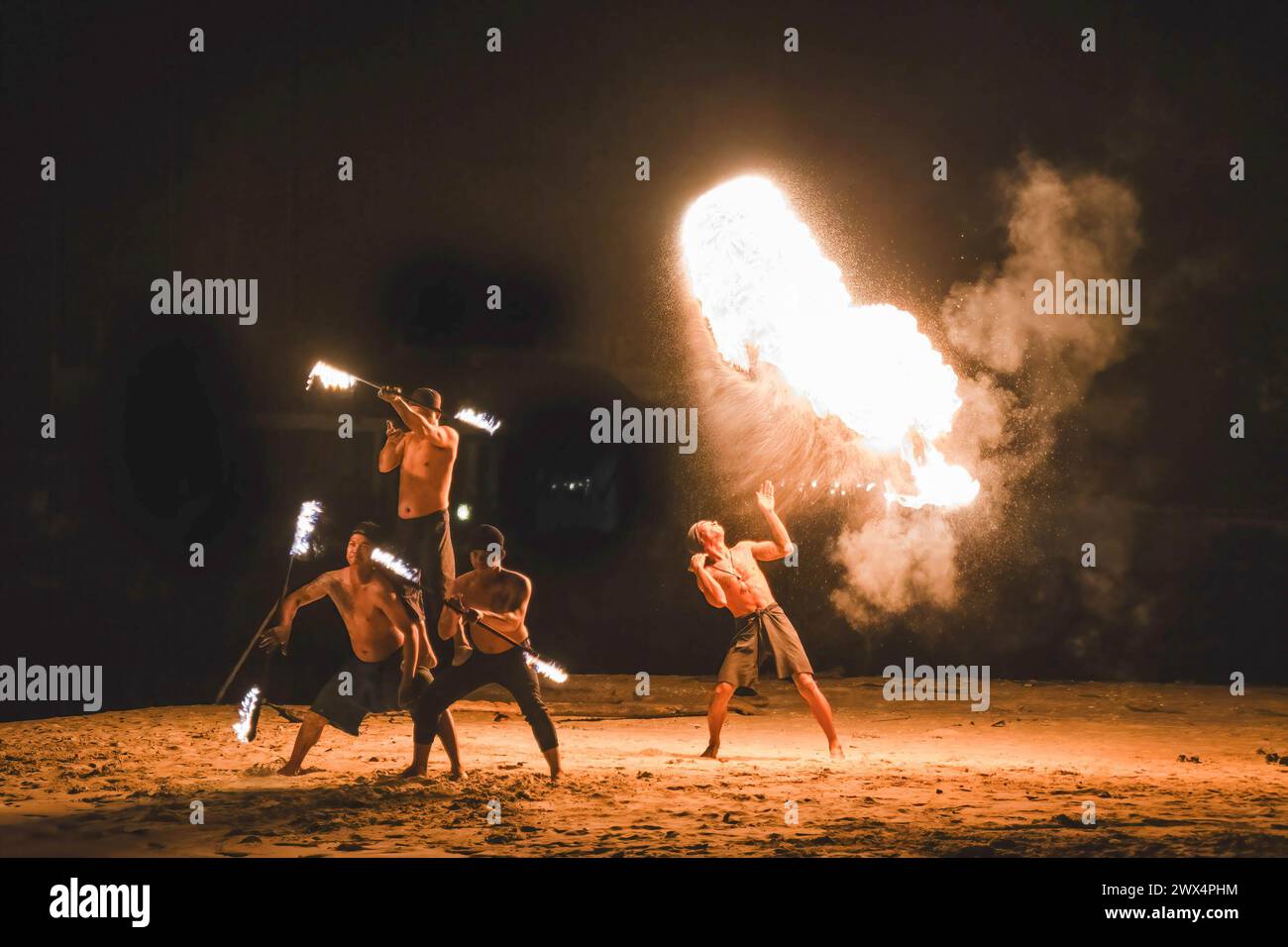 A group of fire dancers juggle and spit fire, on a sandy part of a ...