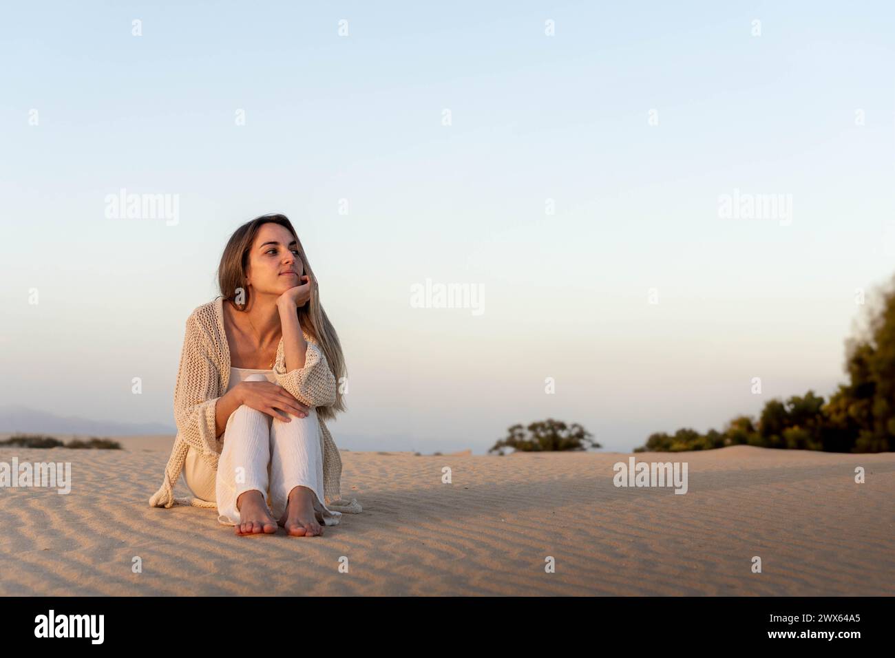 A woman sits peacefully on a sandy dune, her gaze thoughtfully directed ...