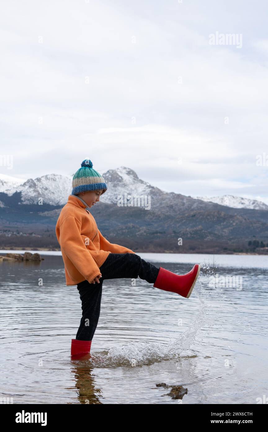 Boy playing with water in a lake wearing wellies with snowy mountains ...