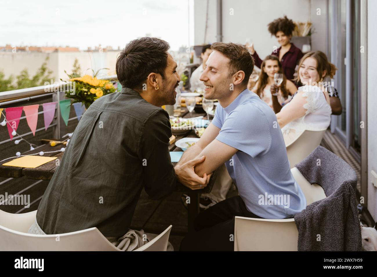 Romantic gay couple looking at each other while sitting with friends at dinner party in balcony ...