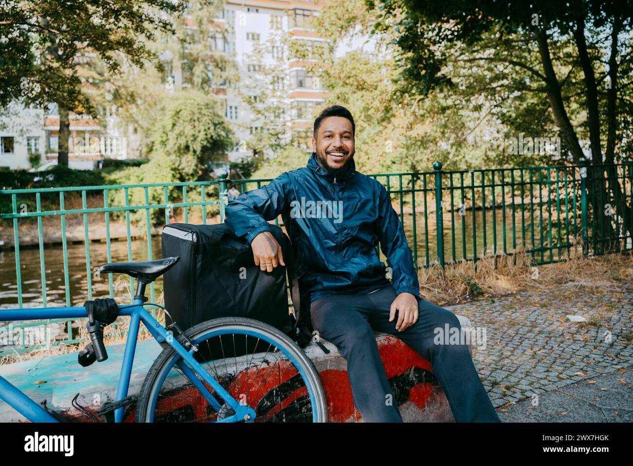 Portrait of happy male food delivery person sitting on bench by bicycle ...