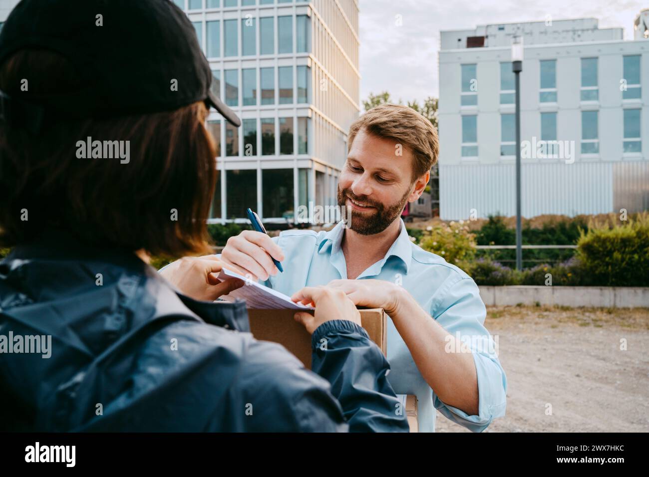 Smiling man signing on document while receiving parcel from delivery ...