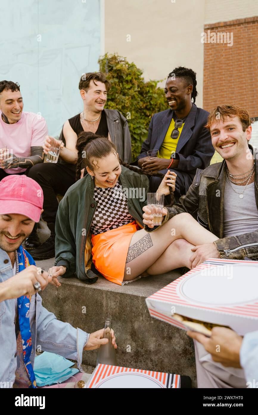 Happy non-binary friends celebrating with food and drinks while sitting ...