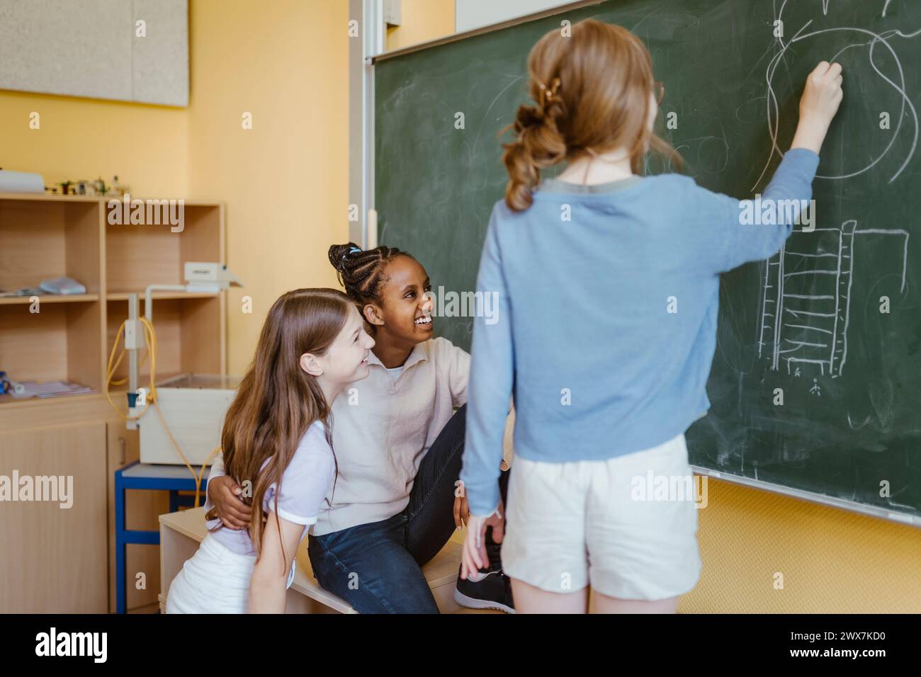 Girls having fun with female friend drawing on chalkboard in classroom ...
