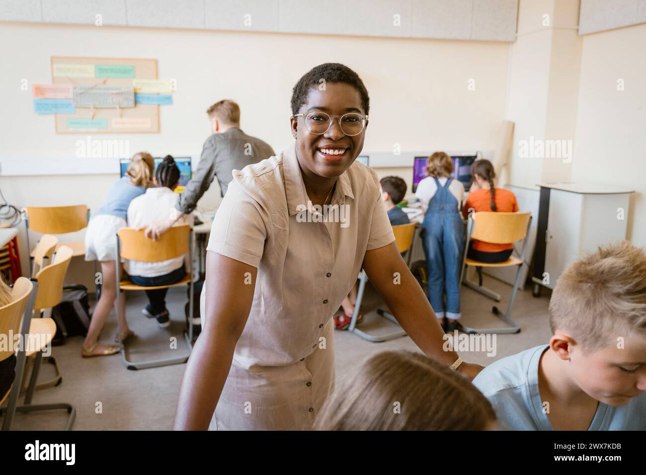 Portrait of female teacher leaning on chairs with students in computer ...