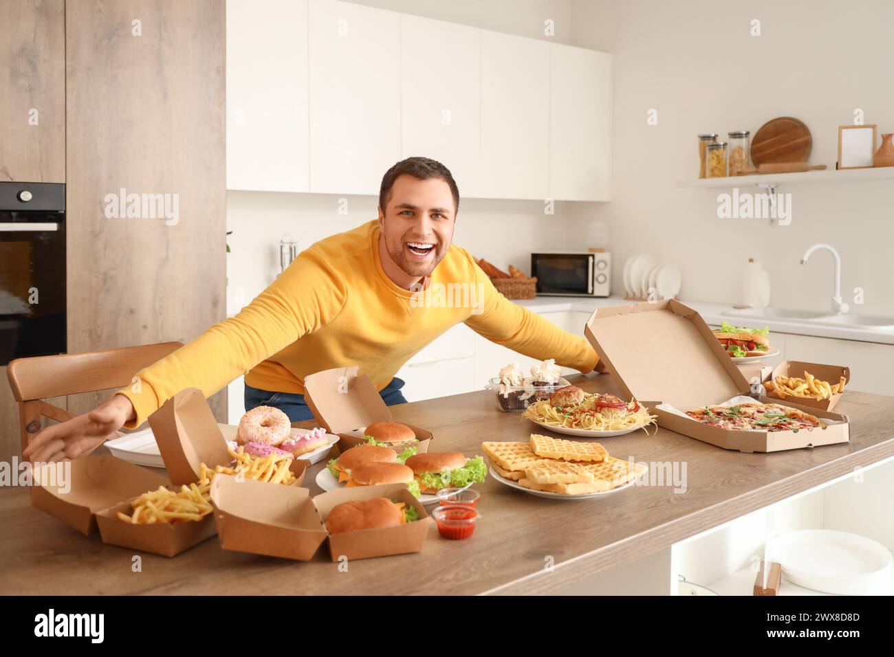 Young man at table full of unhealthy food in kitchen. Overeating ...