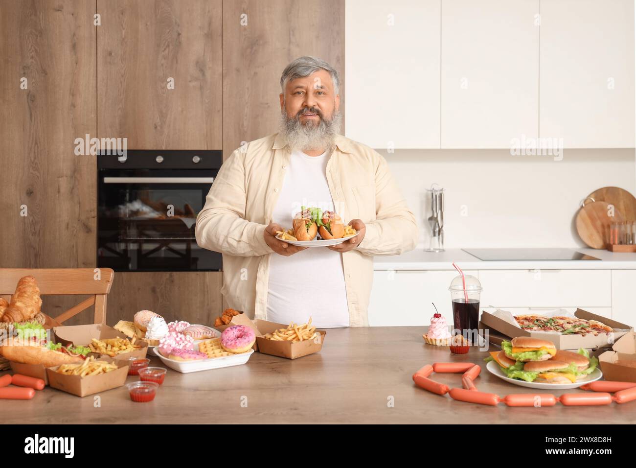 Overweight mature man at table full of unhealthy food in kitchen ...