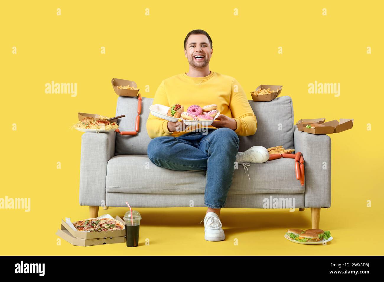 Young man with a lot of unhealthy food sitting on sofa against yellow ...