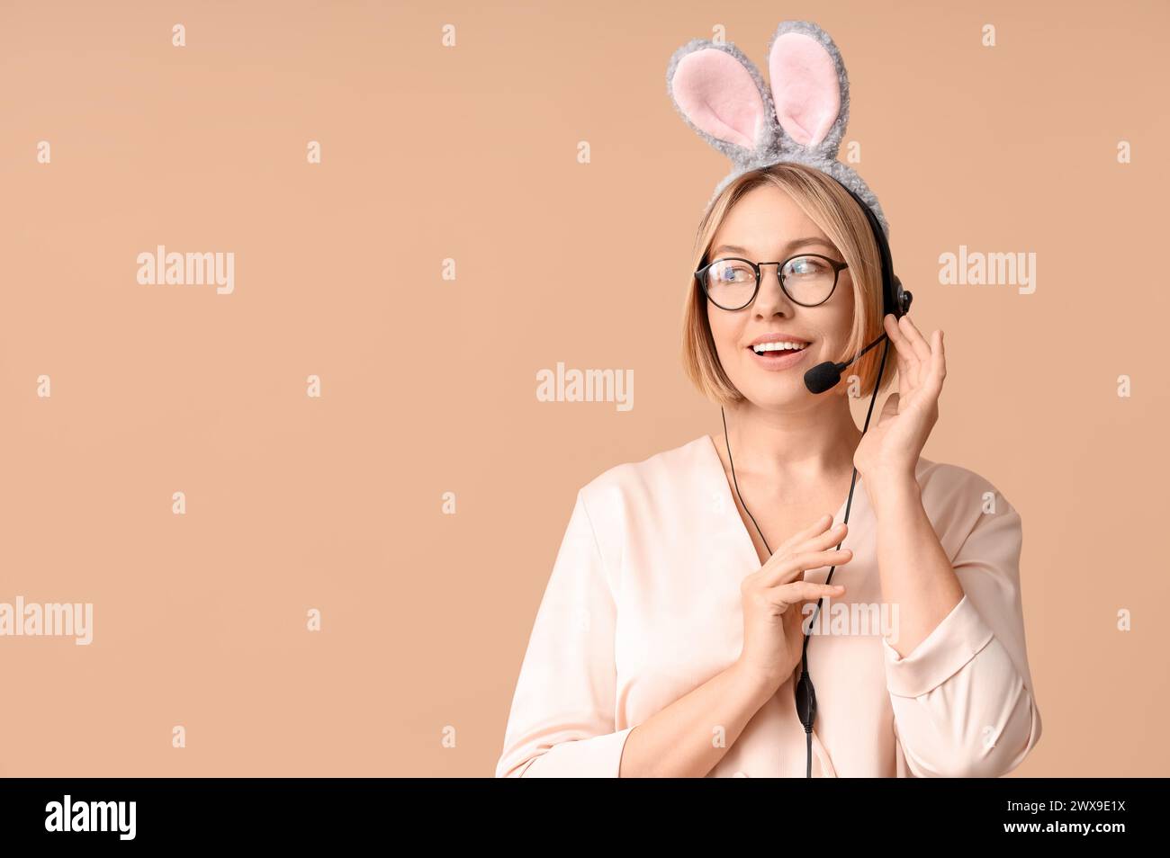 Happy adult female operator in bunny ears with headset on beige ...