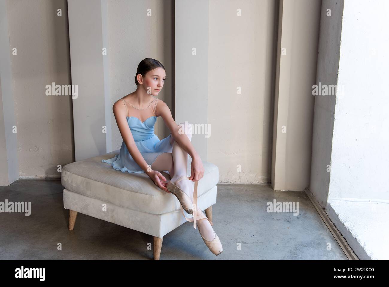 Young serene ballet dancer girl sits on ottoman gracefully securing ...