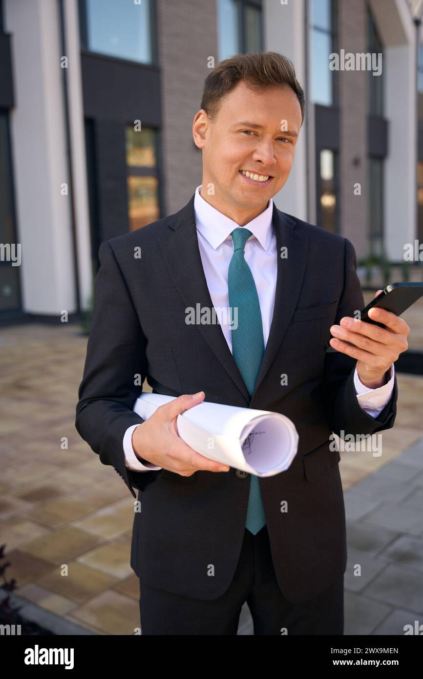 Happy realtor standing with smartphone and building plan in hands Stock ...