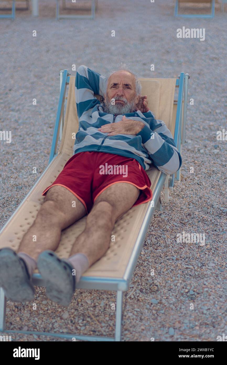 An old man is lying on a sun lounger on the beach. An elderly gray ...