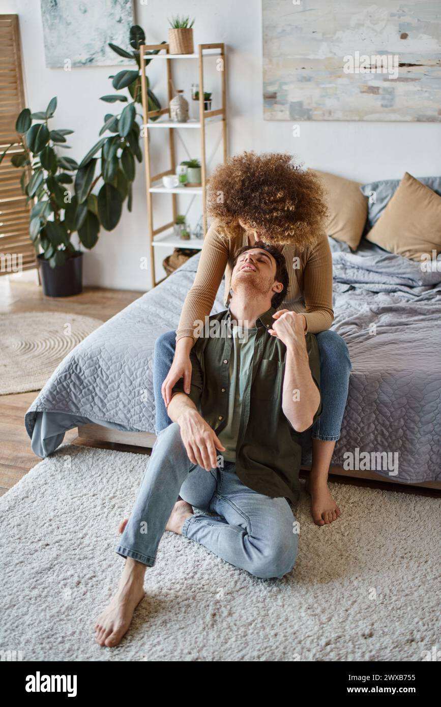 curly young woman hugging her boyfriend from behind sitting on bed ...