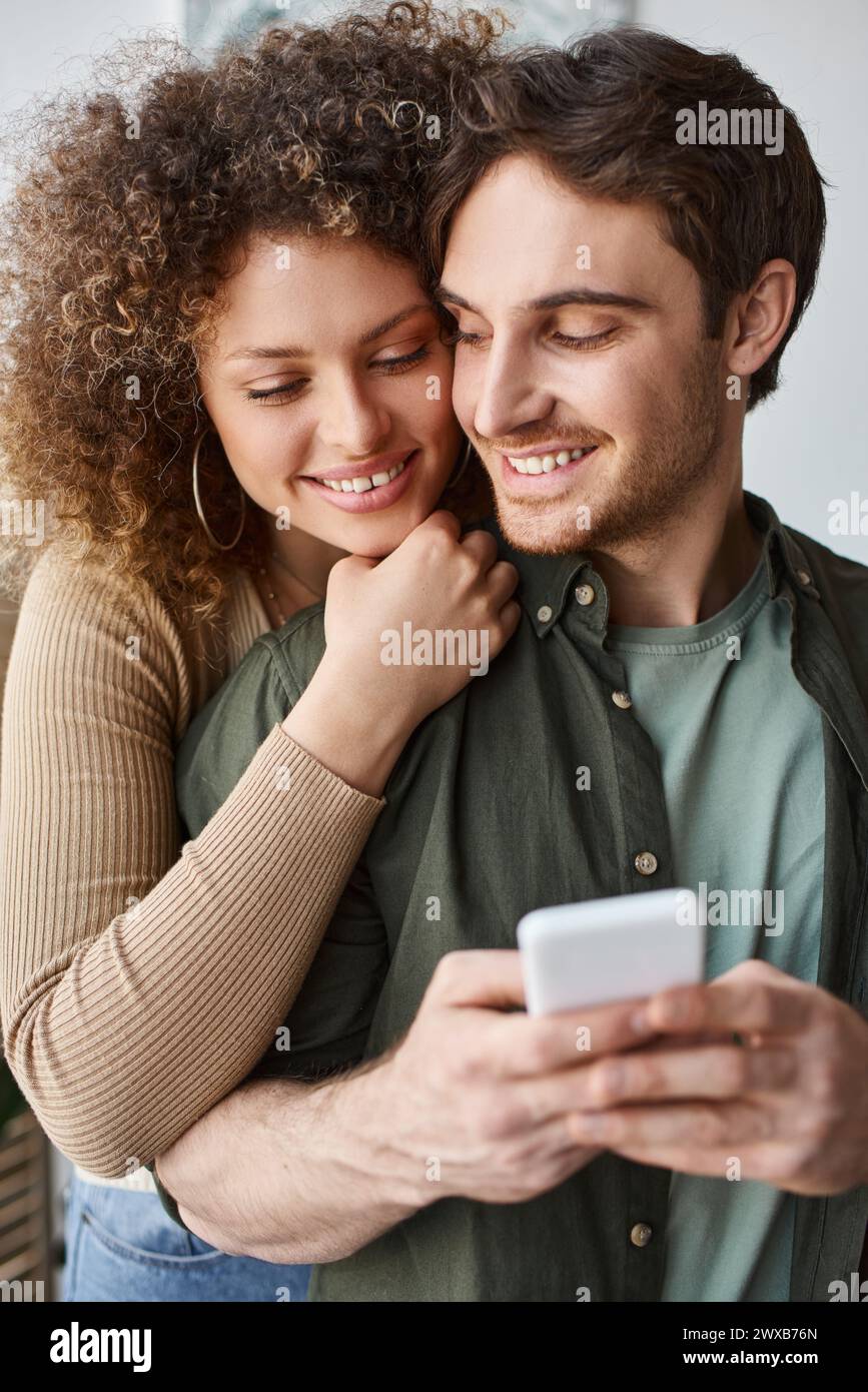 curly young woman kissing her brunette boyfriend, guy holding phone ...