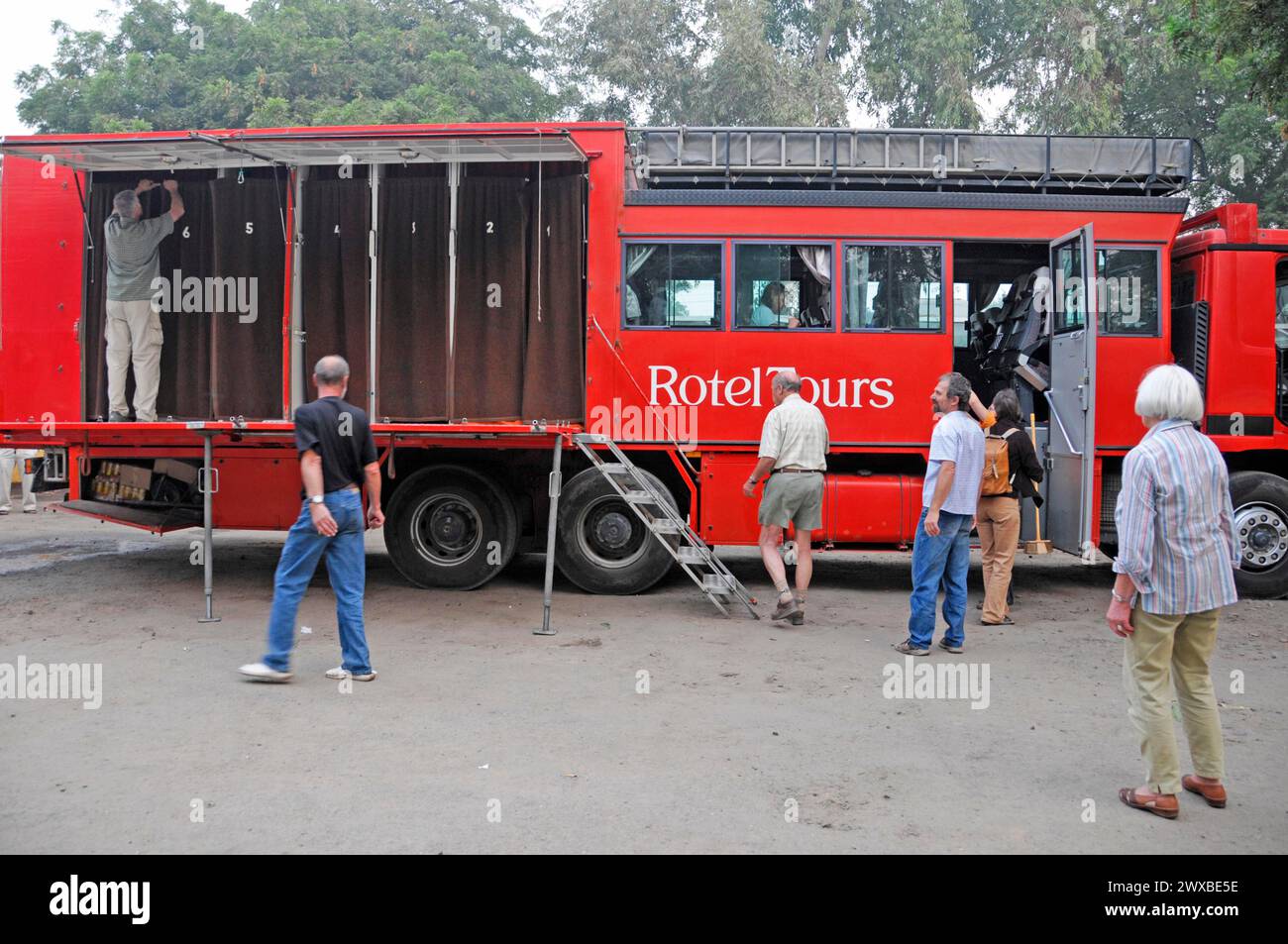 Travellers stand around a red Rotel Tours bus, some get on, Jaipur ...