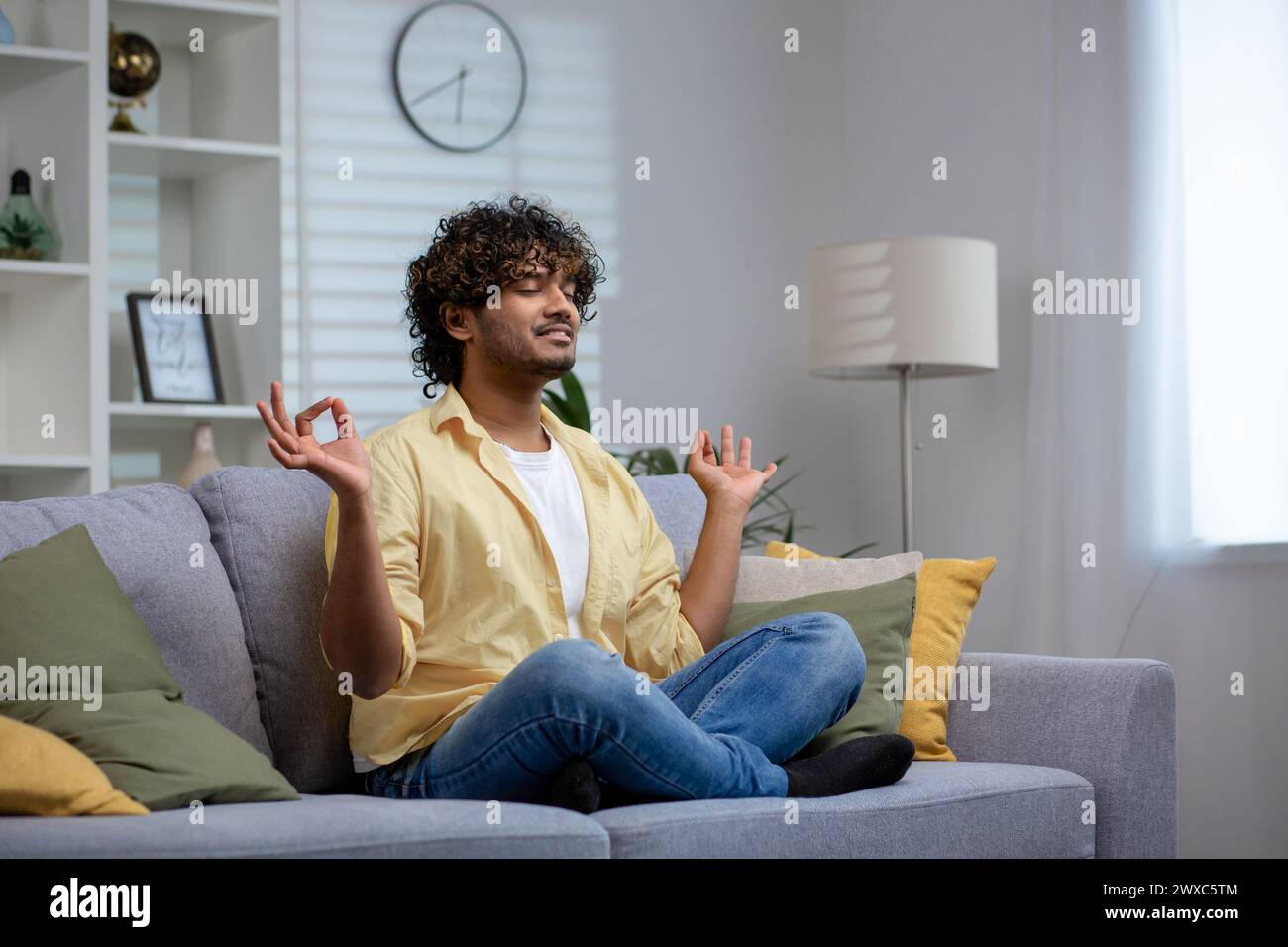 Peaceful Indian man with curly hair meditating in lotus pose on a sofa ...