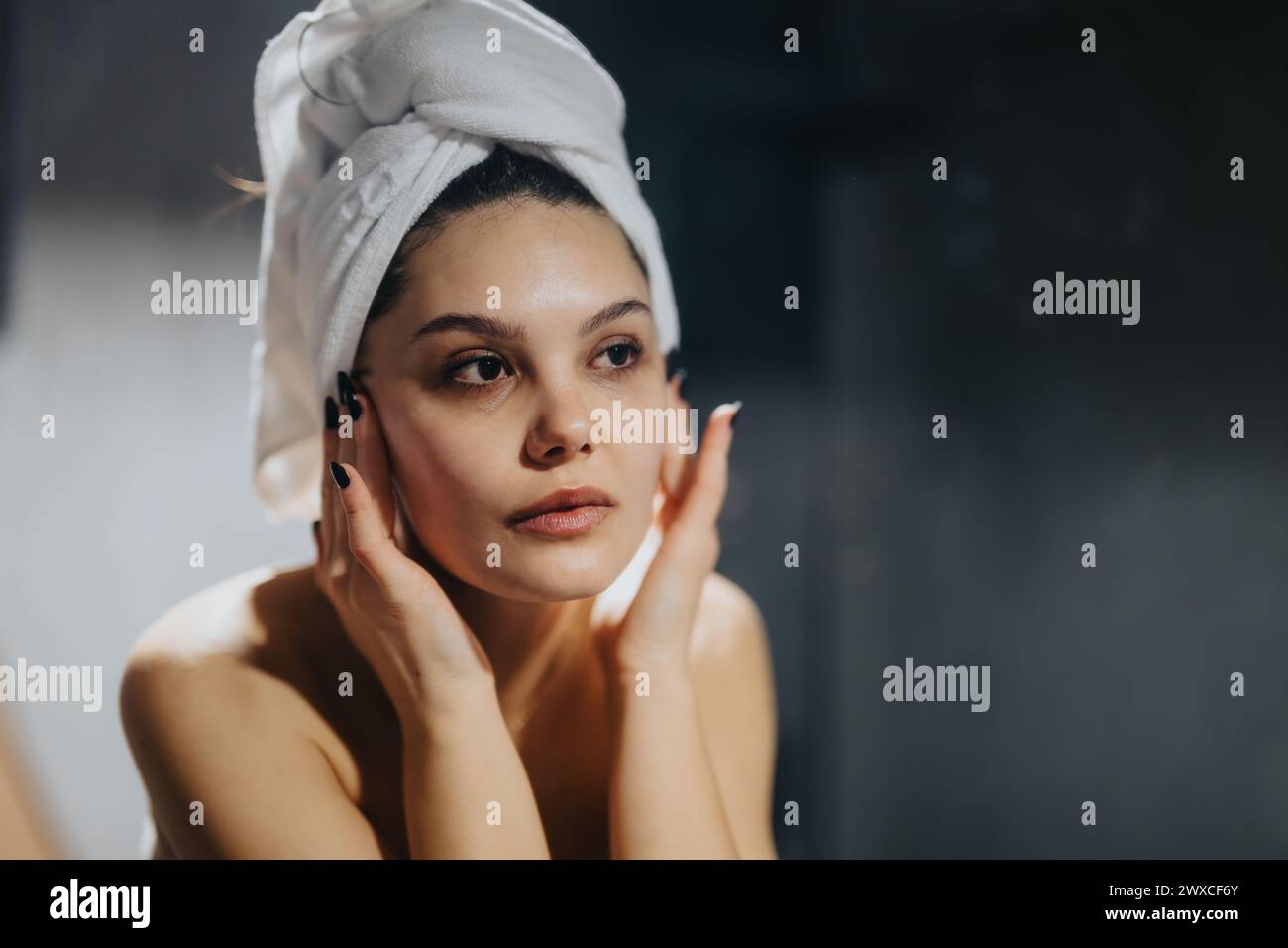 Young woman with a towel on her head checking her face in the mirror ...
