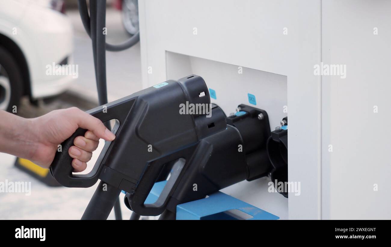 Hand holding EV charging plug in front of camera with blurred ...
