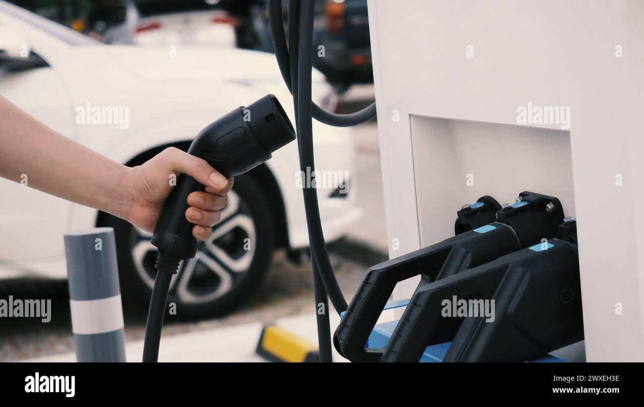 Hand holding EV charging plug in front of camera with blurred ...