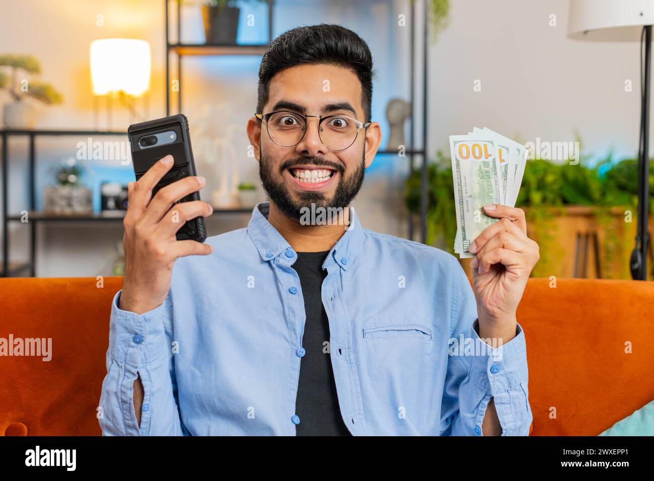 Successful rich Indian man holding smartphone and waving dollar bill money fan sitting on couch ...