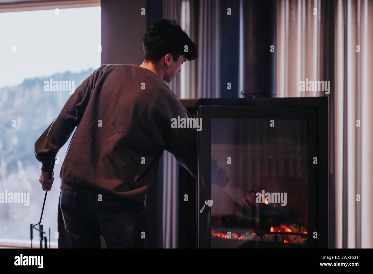 Young male person putting woods at the burning stove, taking care of ...