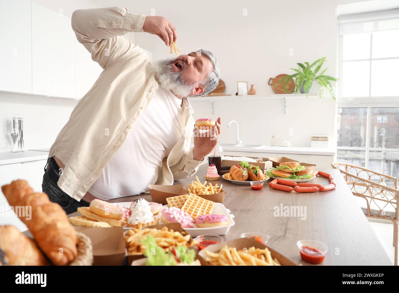 Overweight mature man at table full of unhealthy food in kitchen ...