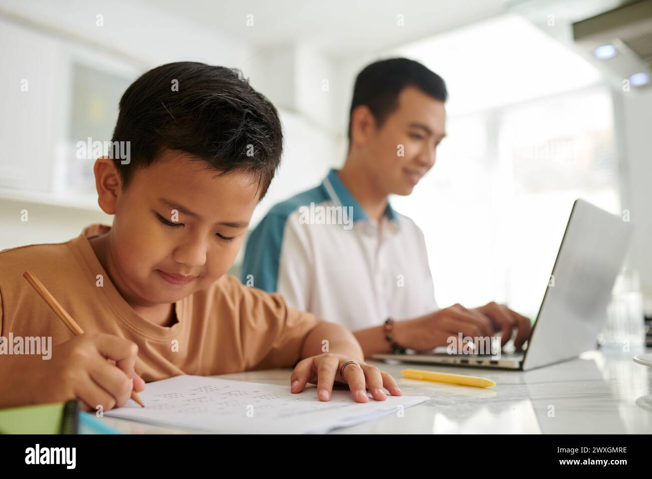 Boy doing homework when sitting next to father working on laptop at ...
