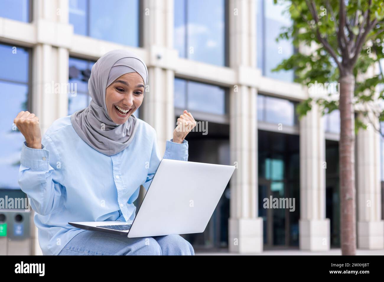 A joyful Muslim woman in a hijab raises her fists in success while ...