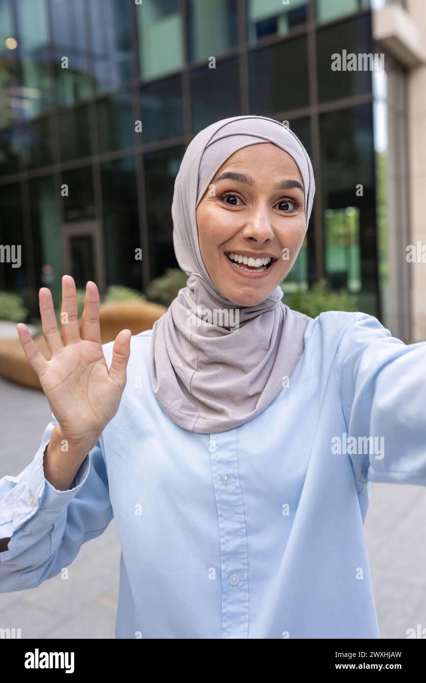 A cheerful woman in a hijab is waving while taking a vertical selfie, seemingly on a video call ...
