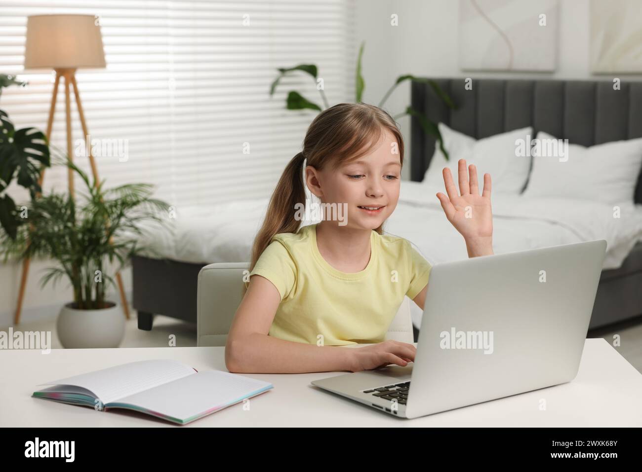 Cute girl waving hello during online lesson via laptop at white table ...
