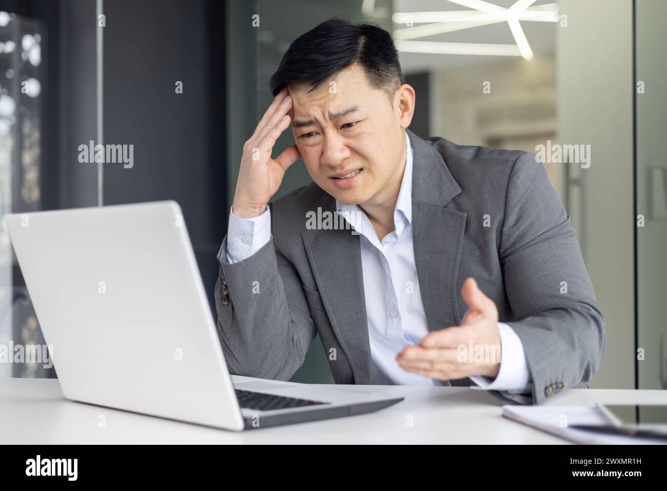 Asian man in business attire looking perplexed at laptop screen while ...